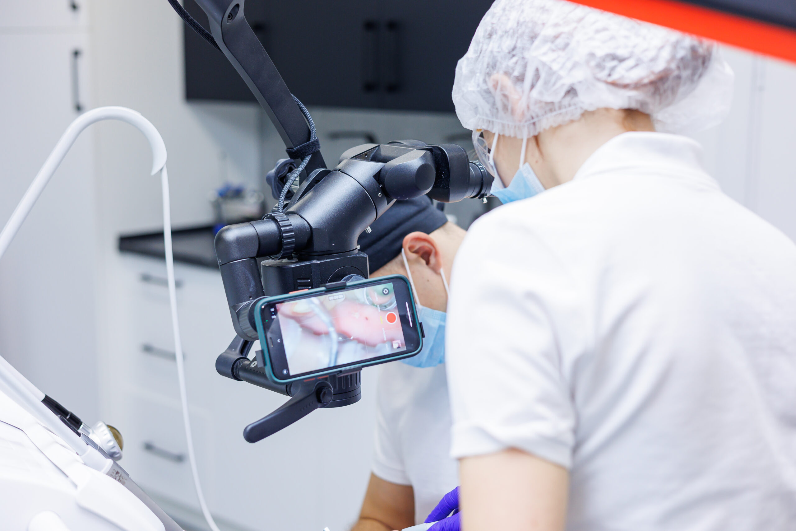 A dentist treats teeth using a dental microscope and tools. The assistant holds a syringe with ...